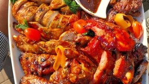 Uma bandeja com carnes grelhadas e legumes, incluindo costelas, asas de frango e camarões, acompanhada de diversos molhos.