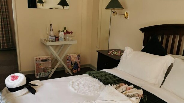 Quarto de hotel com uma cama bem arrumada, uma pequena mesa de apoio com produtos de higiene, uma luminária e um quadro na parede.