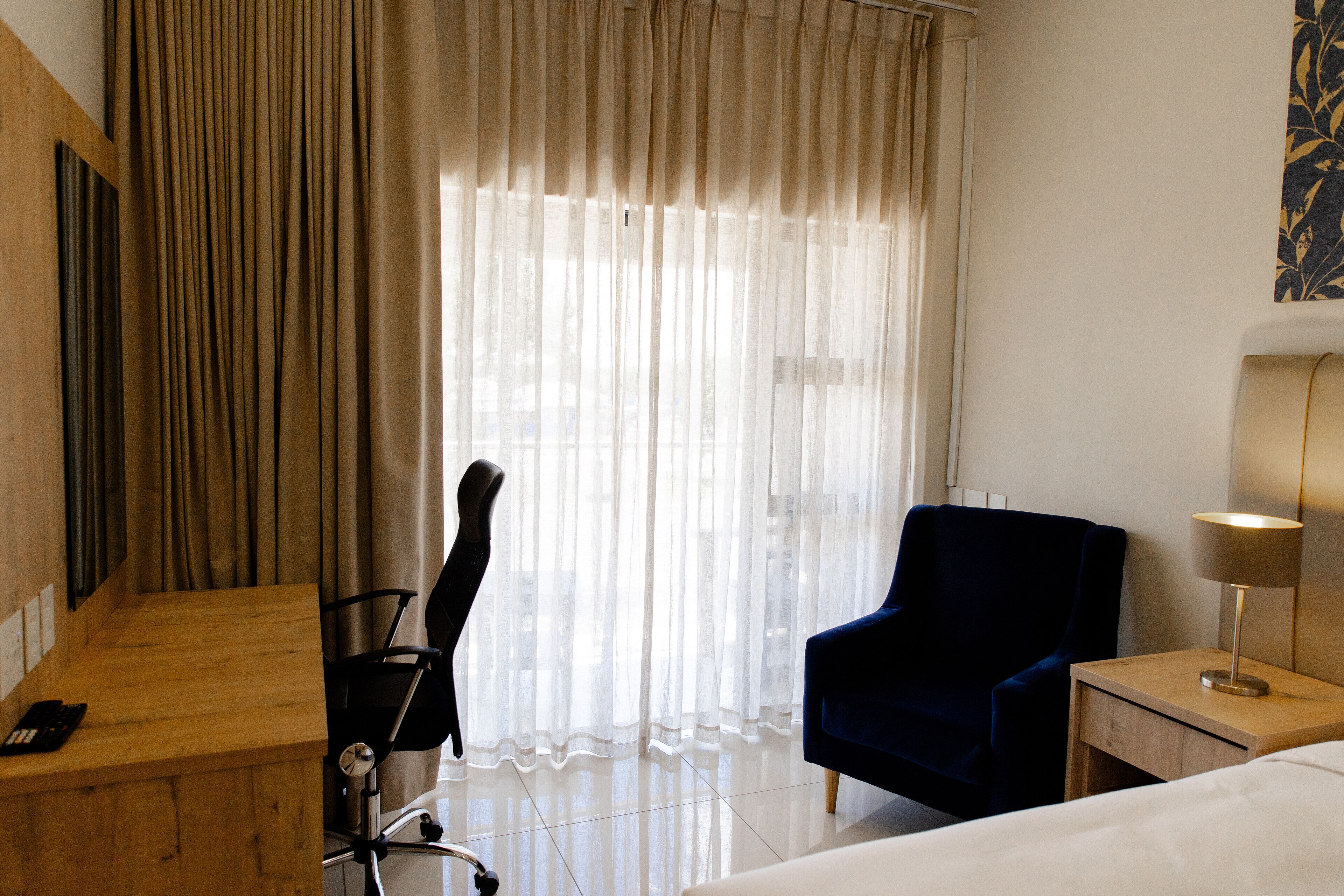 Quarto de hotel com escrivaninha de madeira, cadeira de escritório preta, poltrona azul, mesa de cabeceira com abajur e uma janela grande com cortinas translúcidas.