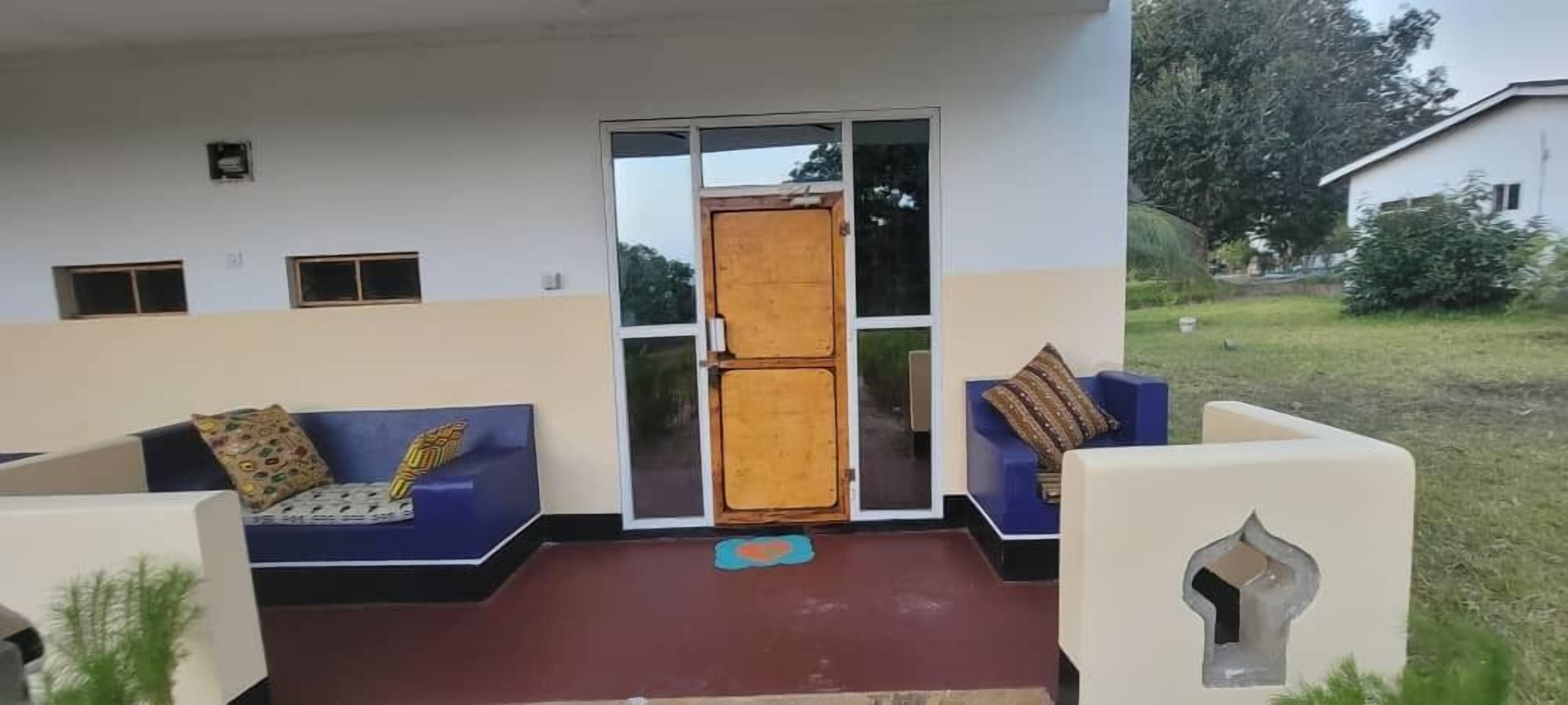 Uma área de estar coberta ao ar livre, com um sofá e almofadas, uma porta de madeira e um pequeno jardim.