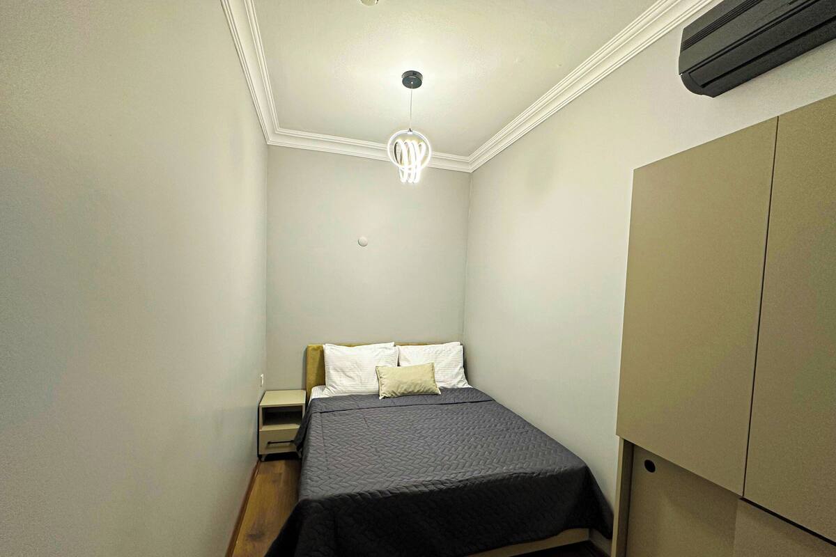 Quarto com cama, mesas de cabeceira, guarda-roupa e luminária no teto.