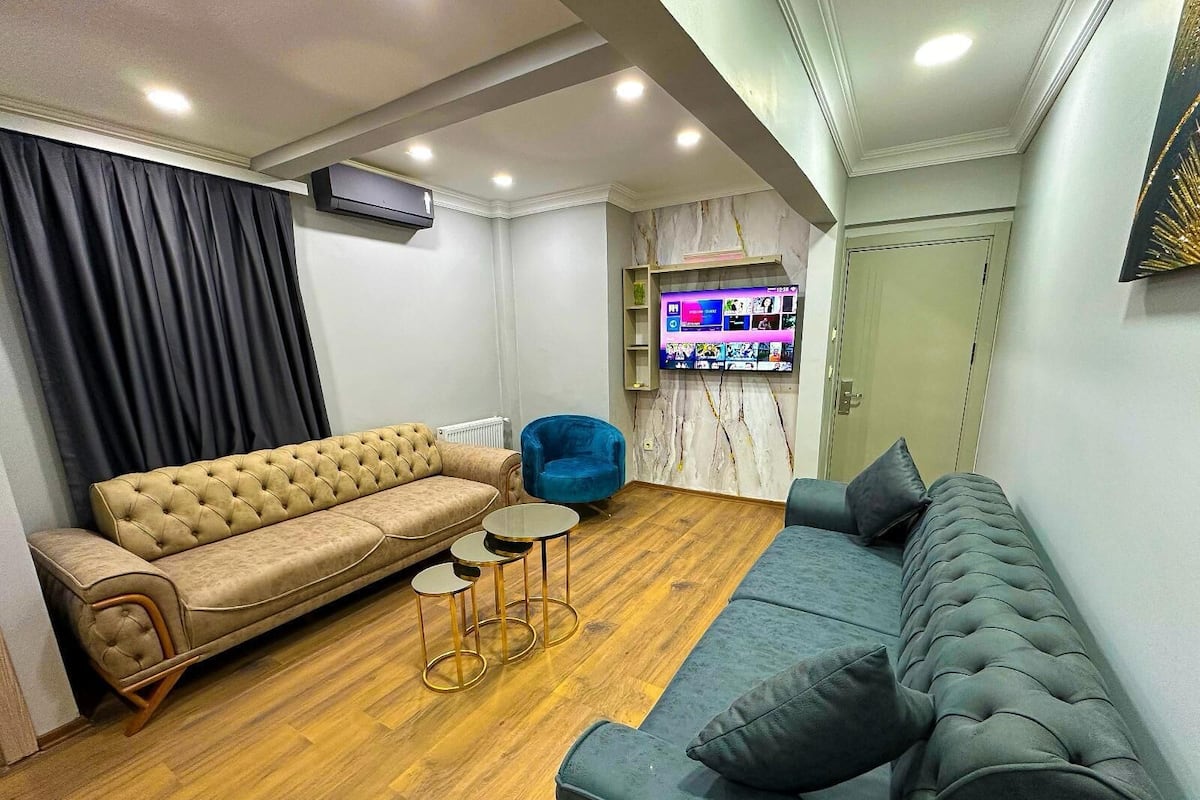 Uma sala de estar moderna com um sofá bege, uma poltrona azul e um sofá verde. Há uma TV de tela plana na parede, uma mesinha de centro e piso de madeira.