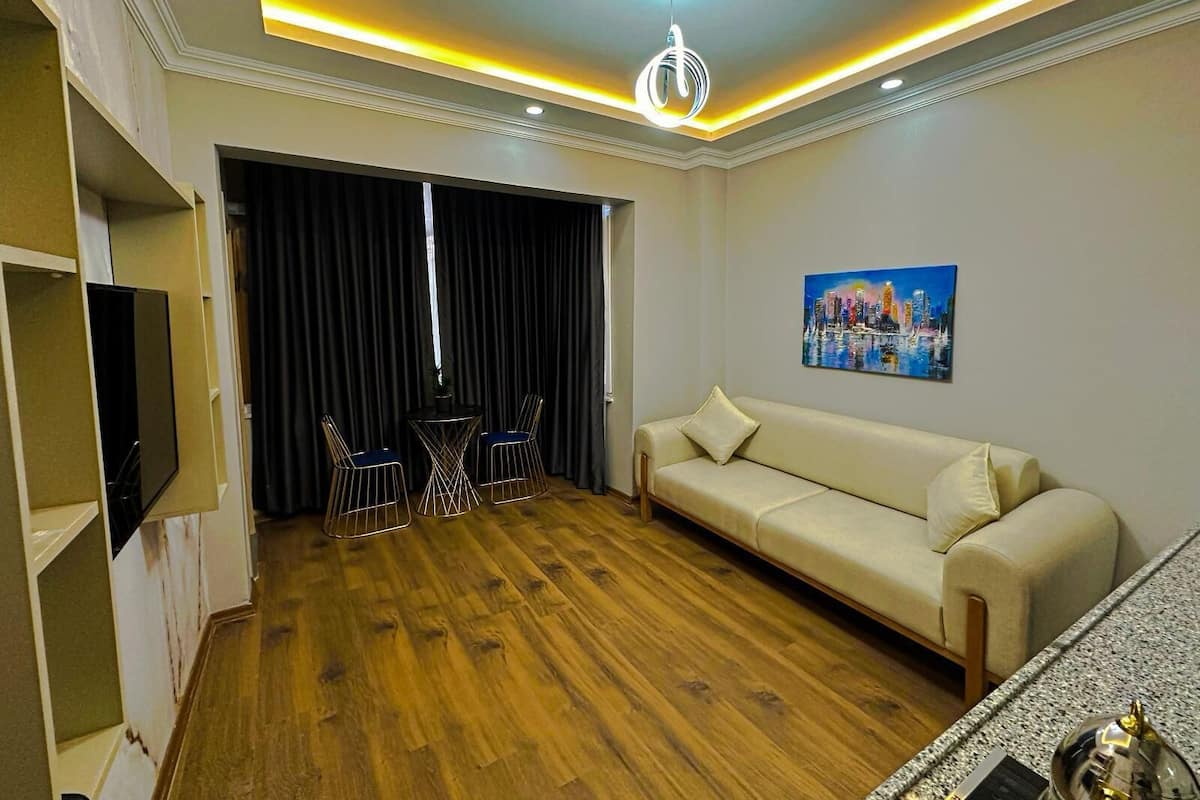 Uma sala de estar moderna com um sofá, uma mesa de jantar com cadeiras, uma televisão de tela plana fixada na parede e uma pintura de uma paisagem urbana.