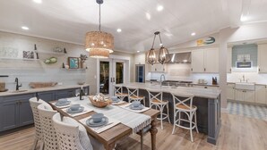 Uma cozinha moderna com ilha central, iluminação pendente e área de jantar com mesa de madeira e cadeiras brancas.