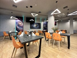 Uma sala de jantar moderna com mesas e cadeiras laranja, com luminárias geométricas no teto e arte nas paredes com tema espacial.