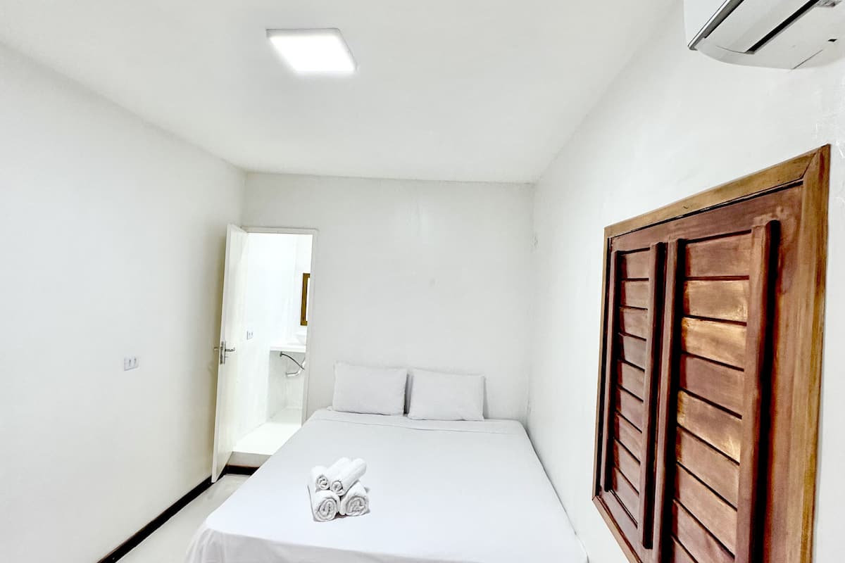 Quarto com uma cama de solteiro, roupa de cama branca, uma porta de madeira e um aparelho de ar condicionado na parede.