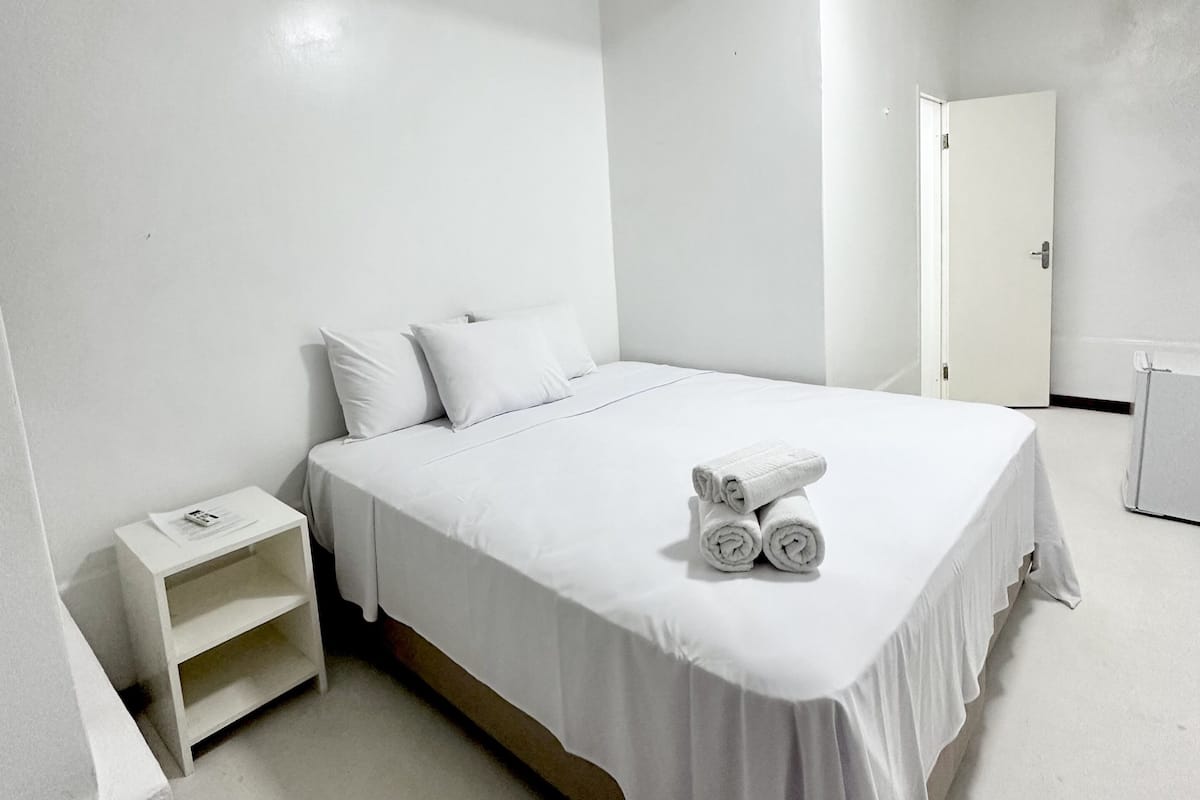 Quarto branco e minimalista, com uma cama grande, uma mesa de cabeceira e uma porta.