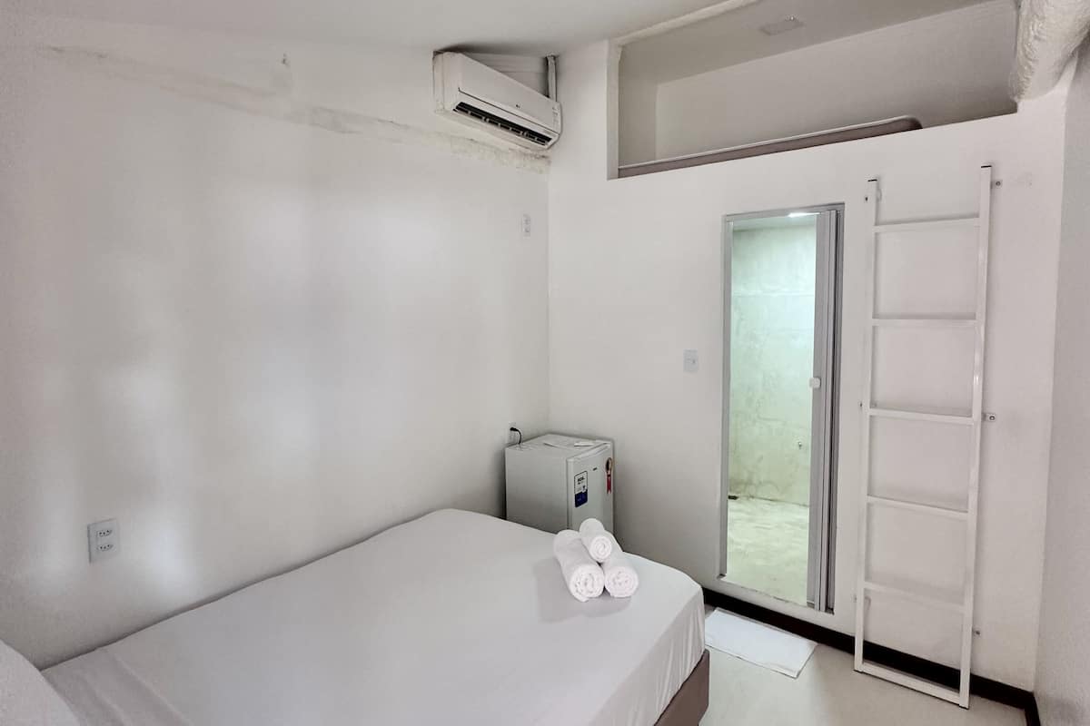 Quarto com cama, ar condicionado, uma mesinha com tomada e porta que dá para uma varanda.