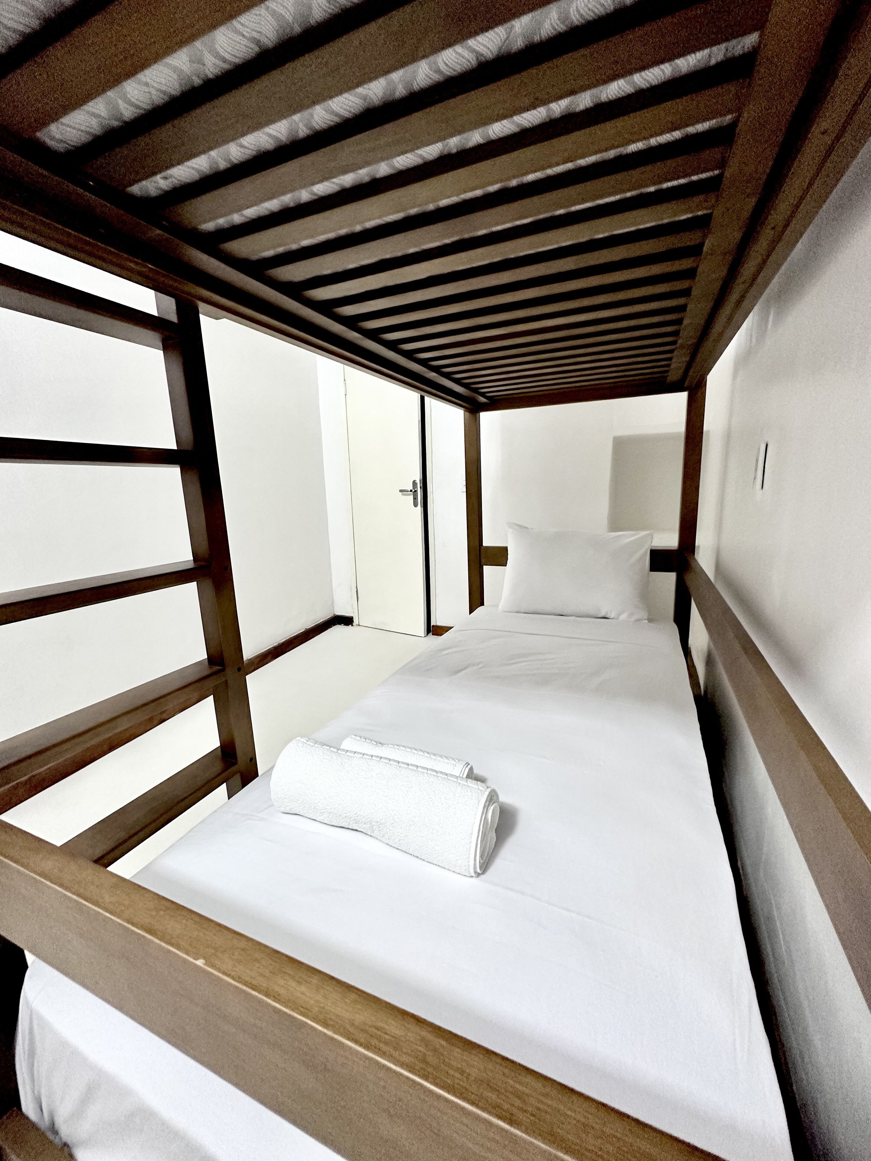 Uma cama de solteiro com estrutura de madeira e roupa de cama branca, uma pequena mesa de cabeceira e uma janela com persianas horizontais.