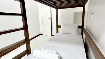 Uma cama de solteiro com estrutura de madeira e roupa de cama branca, uma pequena mesa de cabeceira e uma janela com persianas horizontais.