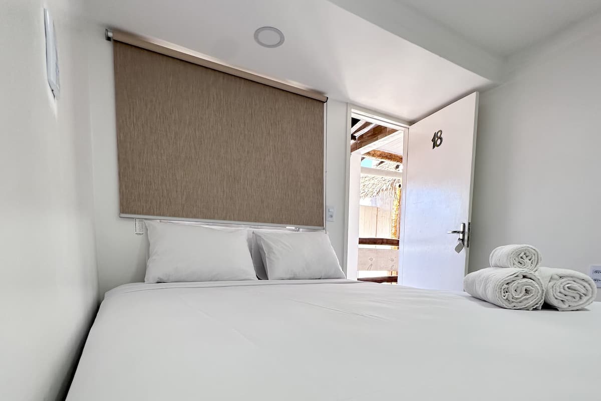 Quarto com cama, cabeceira de madeira, porta com número e toalhas enroladas no chão.