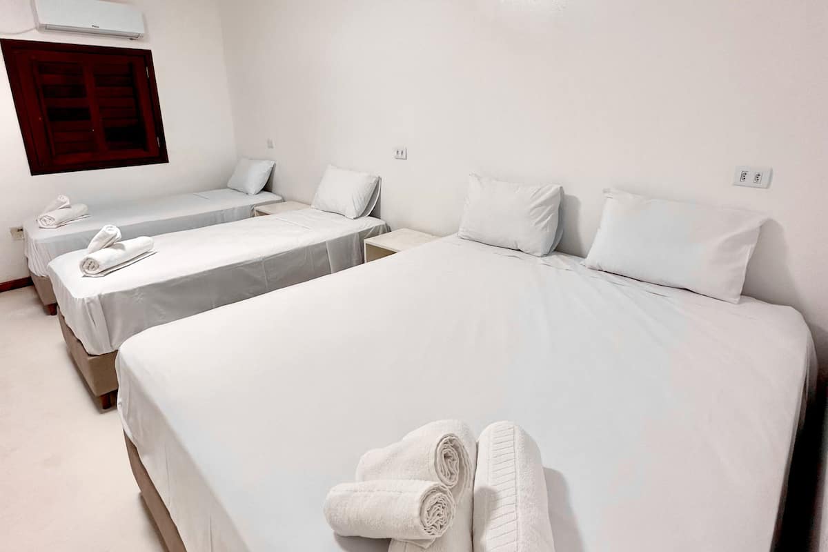 Quarto de hotel com três camas de solteiro e uma cama grande. Cada cama possui um edredom branco e toalhas dobradas.
