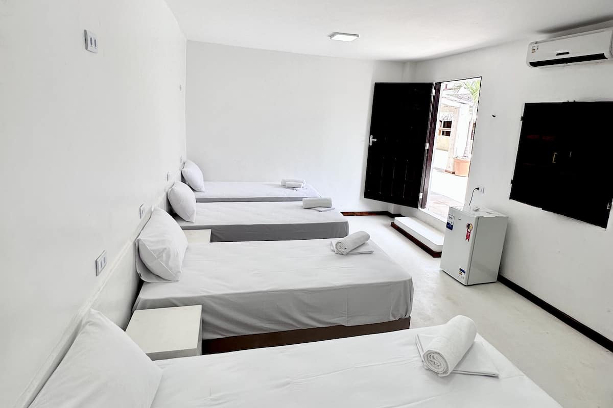 Quarto com quatro camas de solteiro, cada uma com almofada e toalha enrolada. Há um ar condicionado branco na parede, uma porta que dá para o exterior e uma janela com persianas.