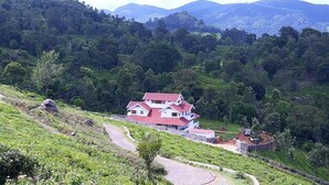Uma casa com telhado vermelho e paredes brancas, rodeada por vegetação exuberante e colinas ao fundo.