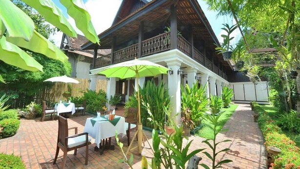 Uma casa de madeira tradicional com varanda, rodeada por vegetação exuberante e um pátio pavimentado com mesas e cadeiras.