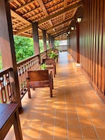 Área externa coberta com bancos de madeira e chão de azulejos, apresentando um corrimão de madeira e plantas em vasos.