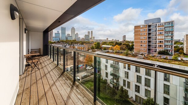 Uma varanda com piso de madeira e guarda-corpo de vidro, com vista para um horizonte urbano com edifícios modernos e céu claro.