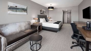 Quarto de hotel com cama, sofá, escrivaninha, cadeira, mesa redonda pequena e televisão.