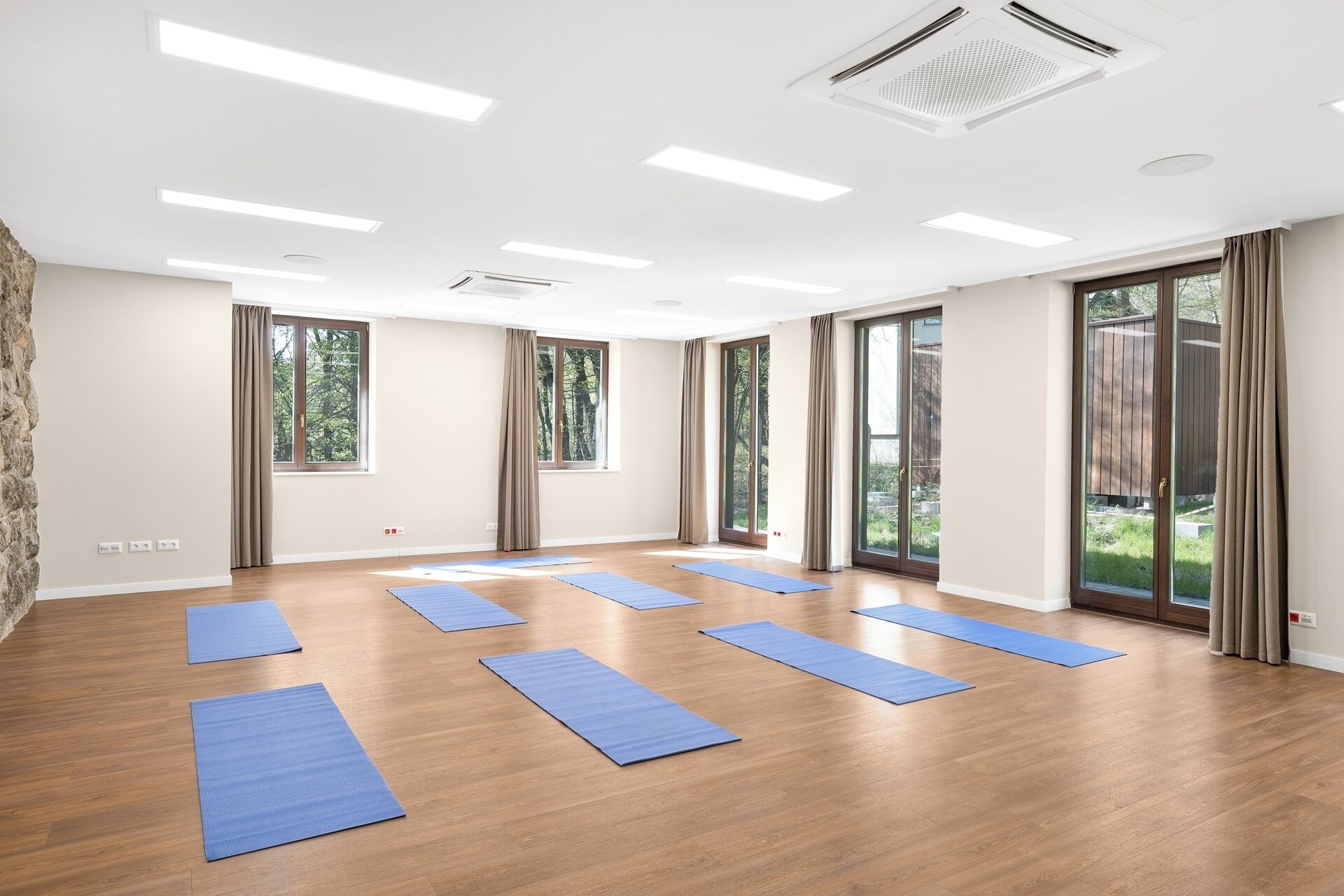 Quarto amplo com assoalho de madeira, grandes janelas e tapetes de yoga azuis dispostos para uso.