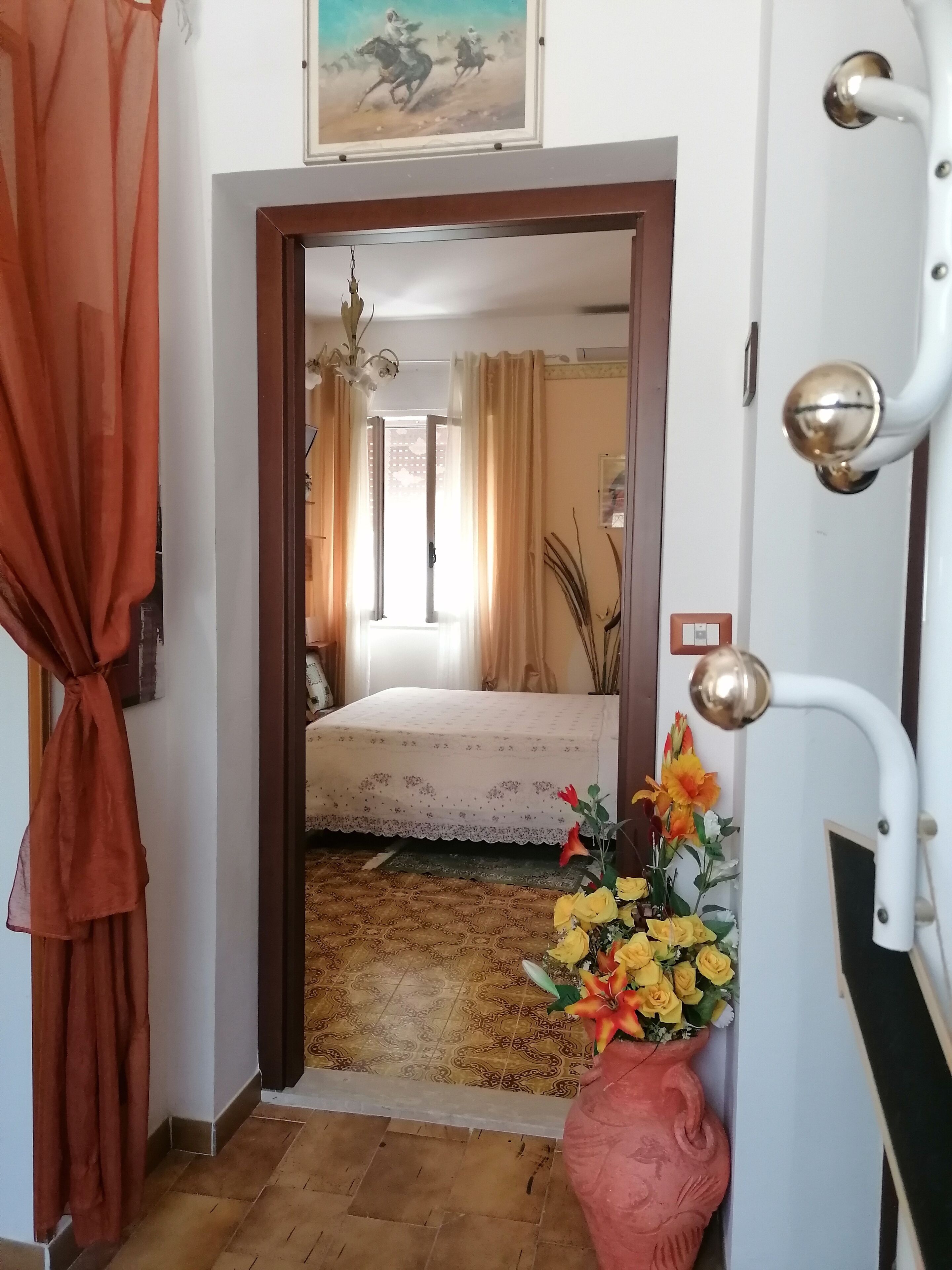 Quarto com cama, porta com puxador de latão, planta em vaso com flores e um quadro na parede.