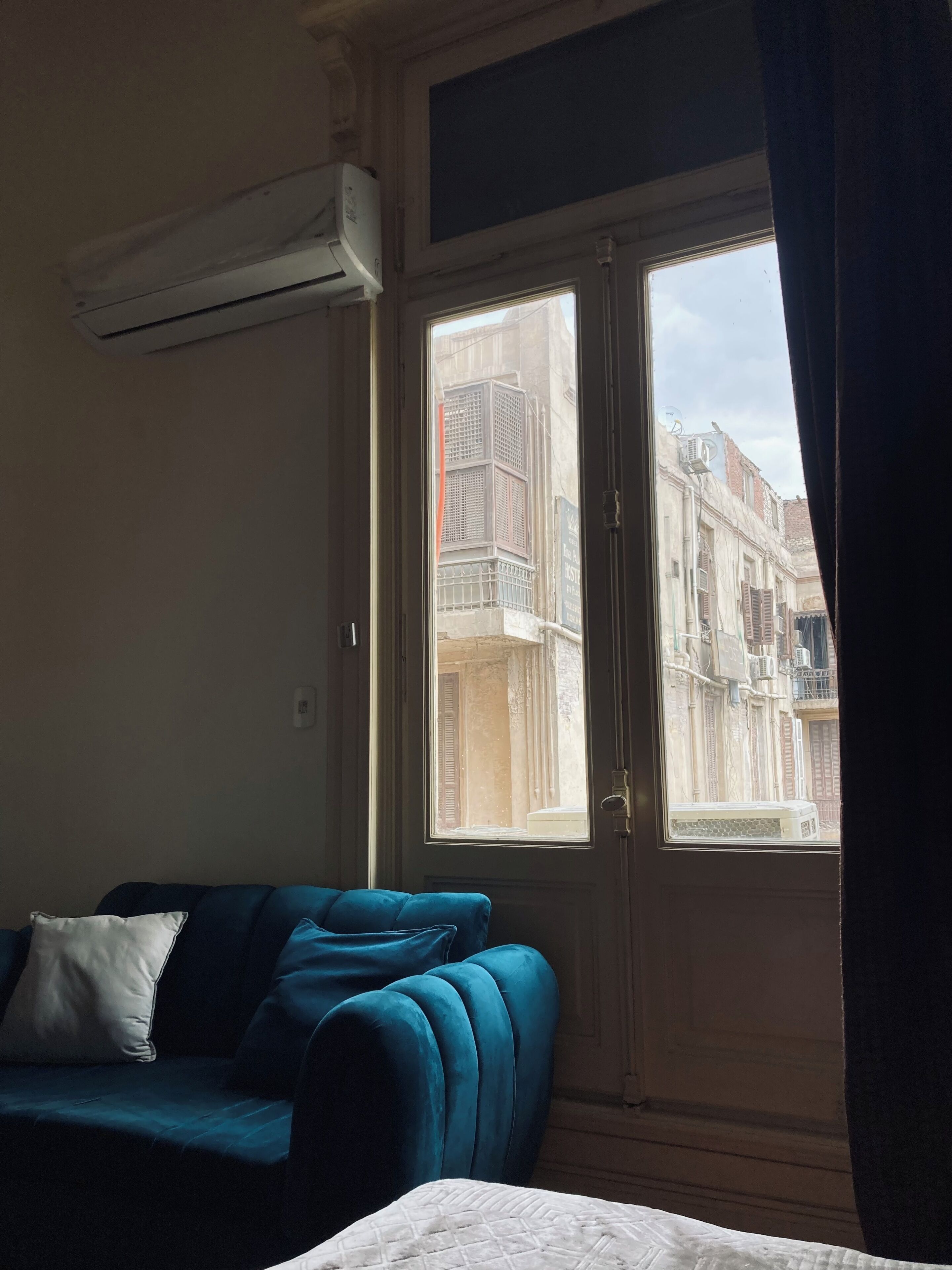 Quarto com um sofá azul, uma janela com vista para edifícios e um aparelho de ar condicionado.