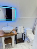 Uma casa de banho com lavatório branco, espelho com iluminação azul, sanita e um porta-toalhas.