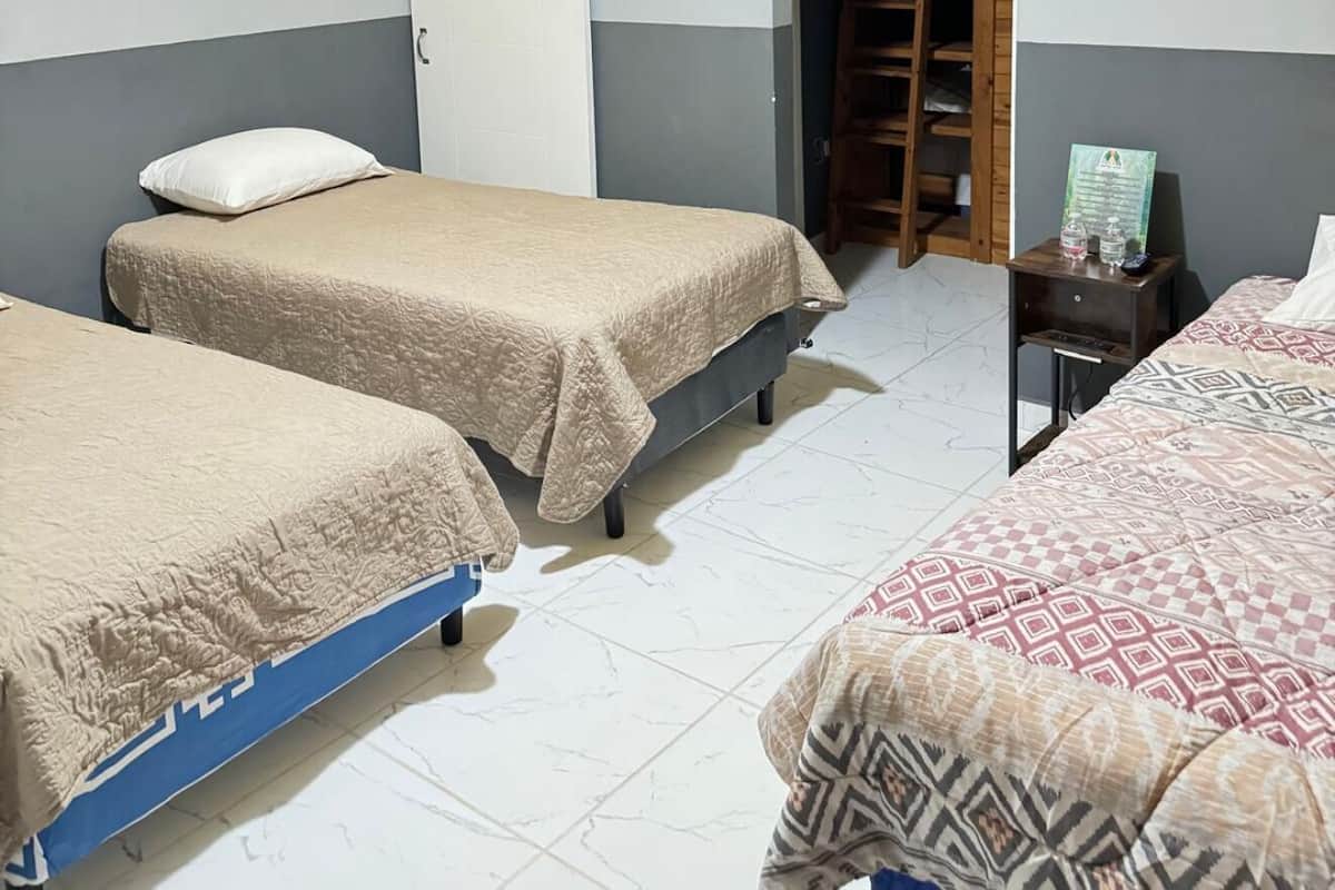 Quarto com duas camas, um ventilador de teto, uma porta e uma pequena mesa com uma cadeira.