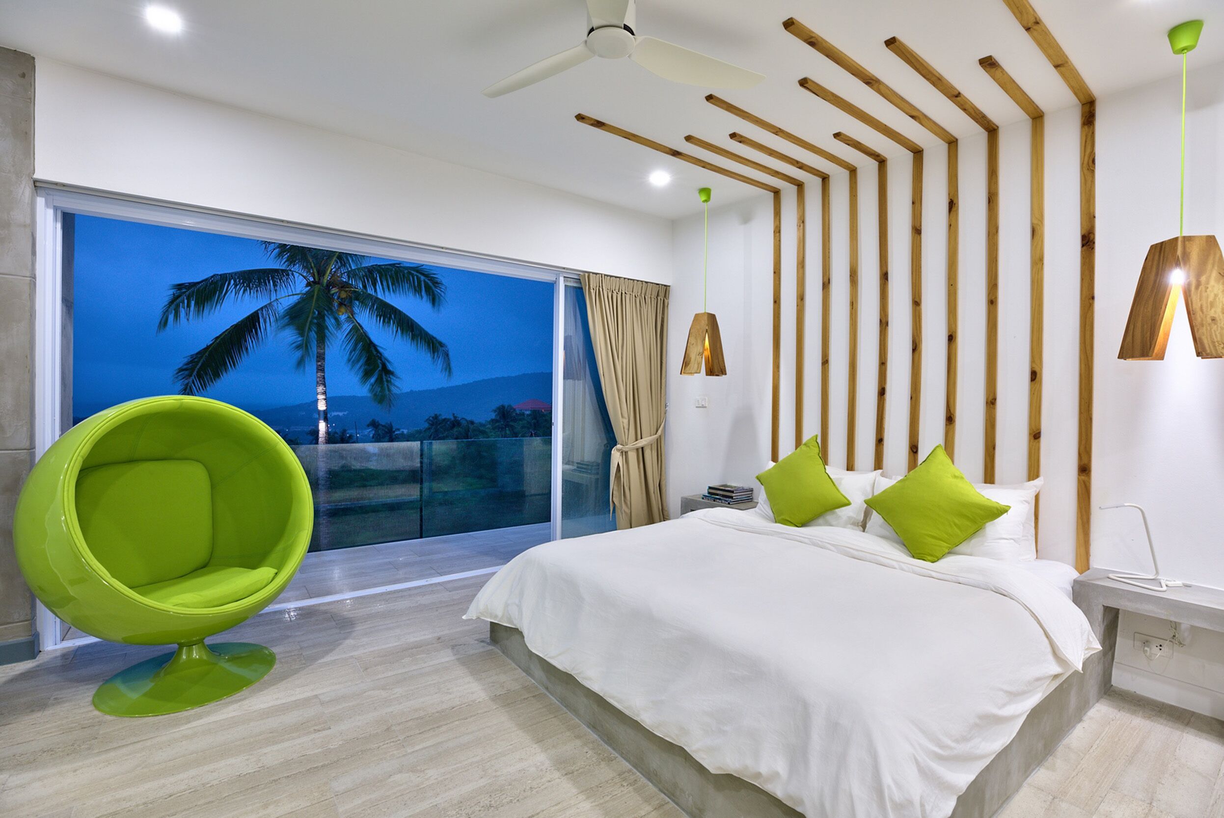 Quarto moderno com uma cama grande, uma cadeira verde vibrante, vigas de madeira no teto e vista para palmeiras pela janela.