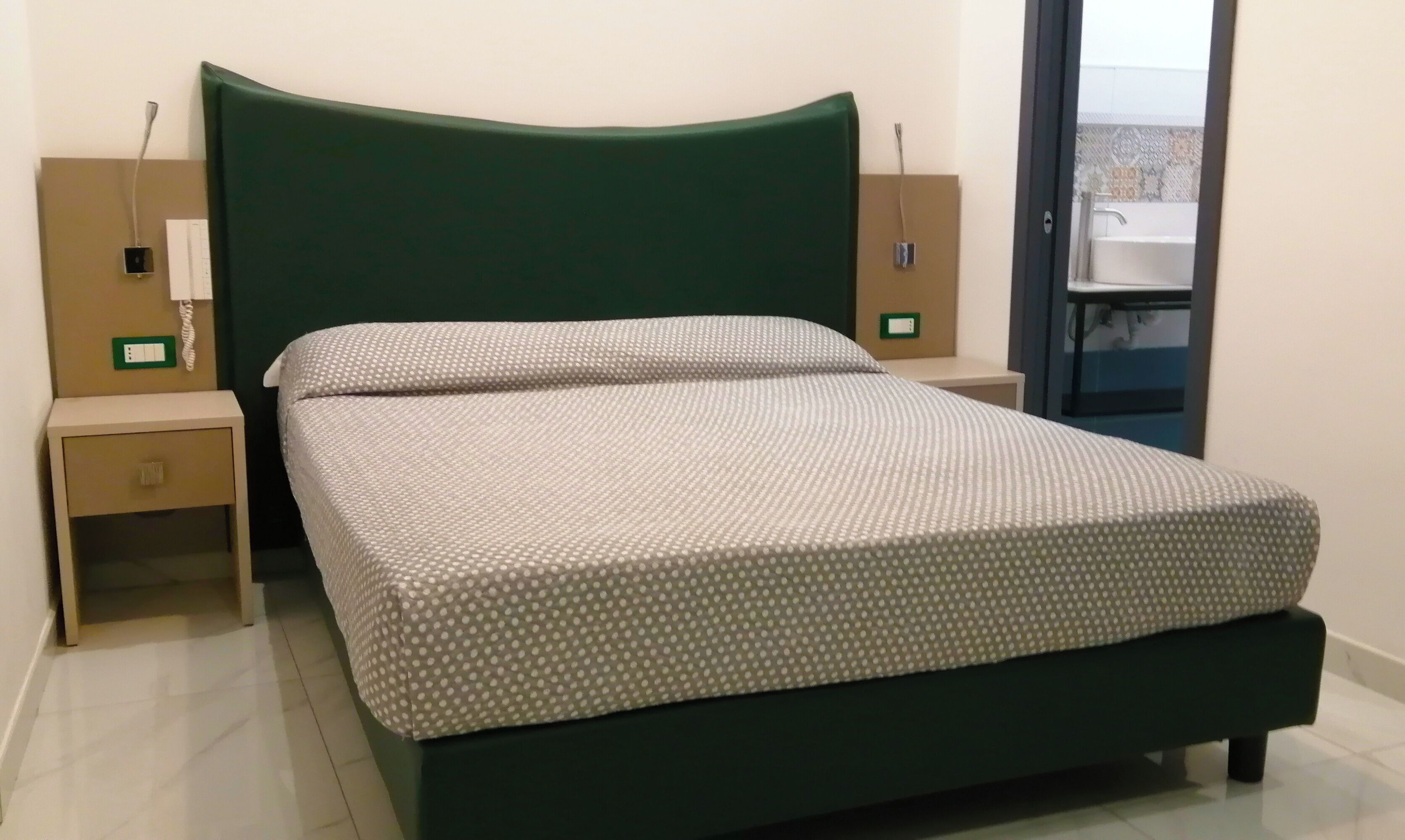 Uma cama com cabeceira verde, edredom branco e bege com bolinhas, e duas mesas de cabeceira com abajures.