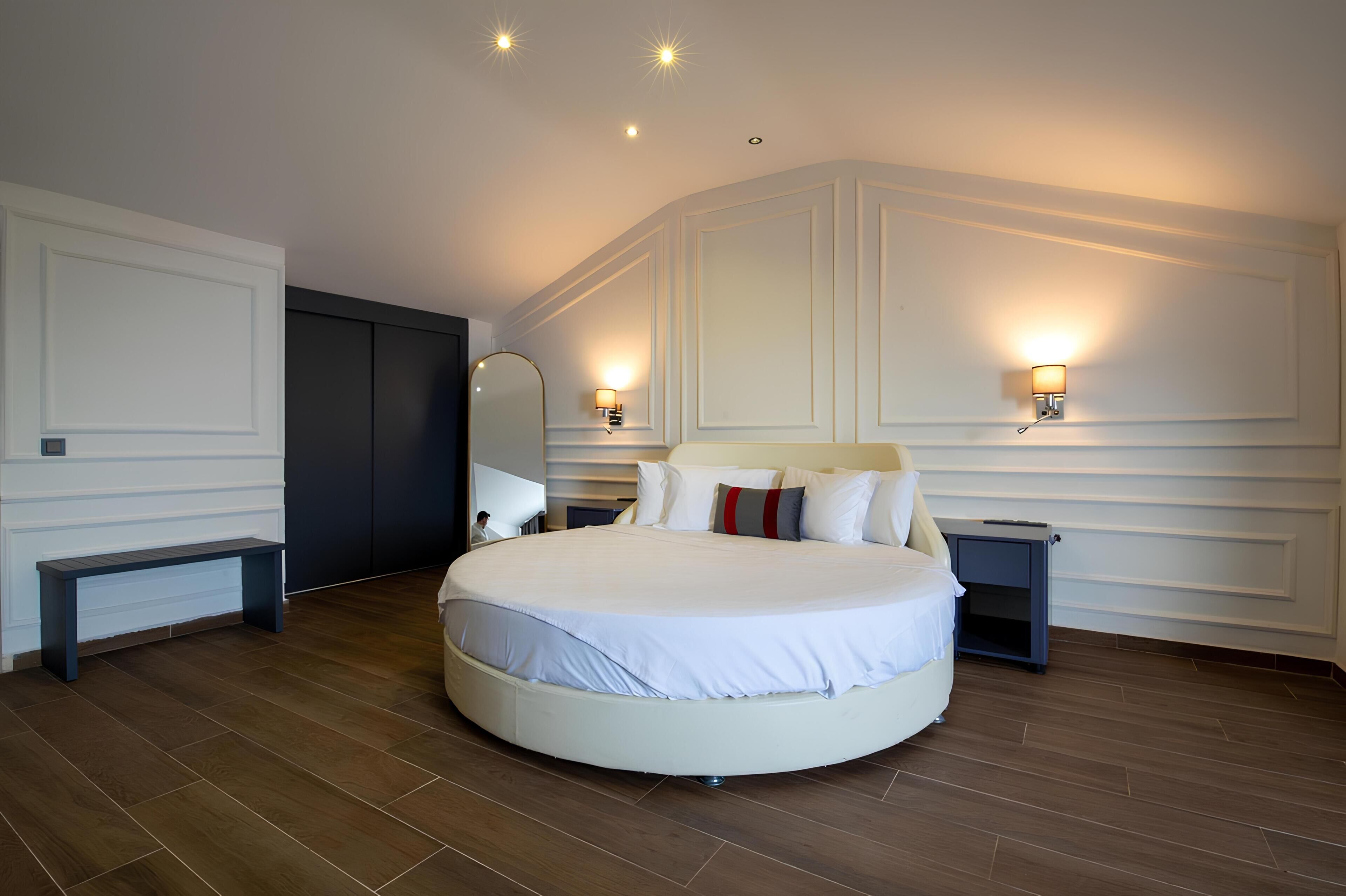 Uma cama redonda grande com roupa de cama branca, duas mesas de cabeceira e luminárias fixadas na parede.