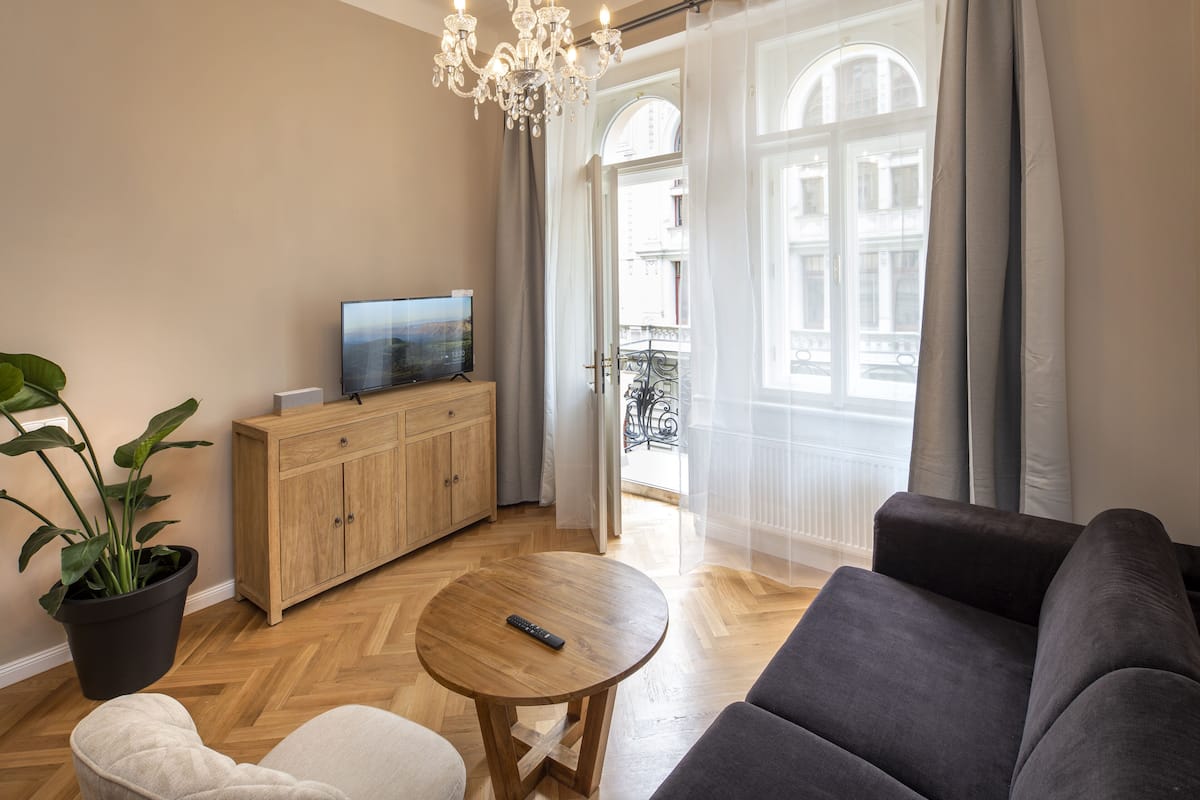 Uma sala de estar com sofá, mesa de centro redonda de madeira, televisão em um armário de madeira, planta em vaso e vista para uma varanda através de cortinas leves.