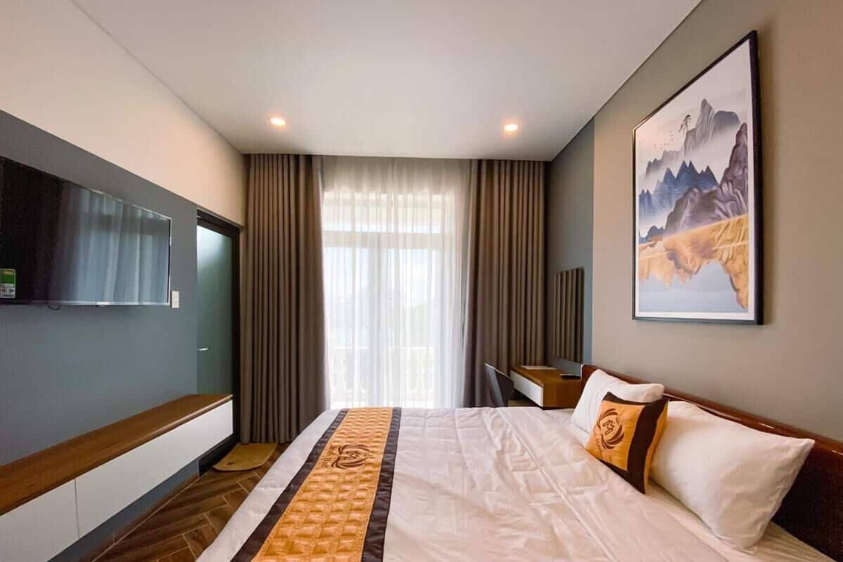 Quarto de hotel com uma cama grande, uma escrivaninha, televisão na parede e um quadro sobre a cama.