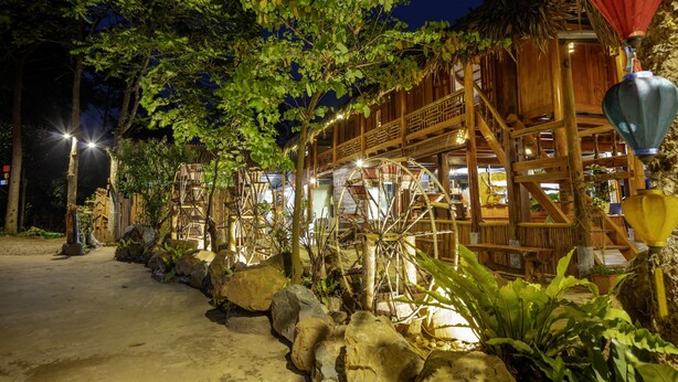 Estrutura de madeira tradicional com telhado de palha, rodeada por pedras e vegetação, iluminada por iluminação suave.