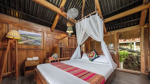 Quarto de madeira com uma cama, mesa de cabeceira, luminária, pintura e janela com vista para o verde.