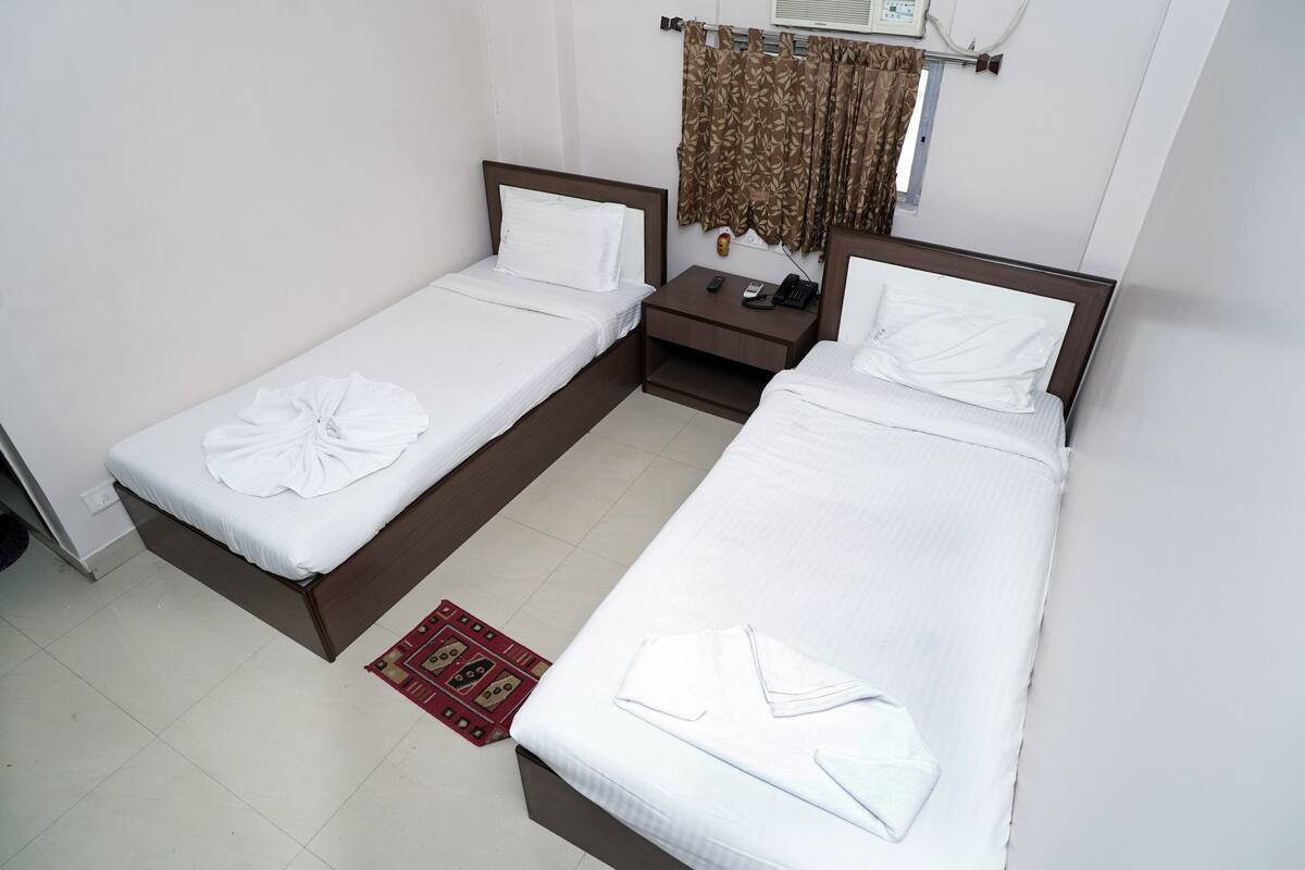 Quarto de hotel com duas camas de solteiro, uma pequena mesa de cabeceira com telefone e luminária, um tapete vermelho estampado e uma janela com cortinas.