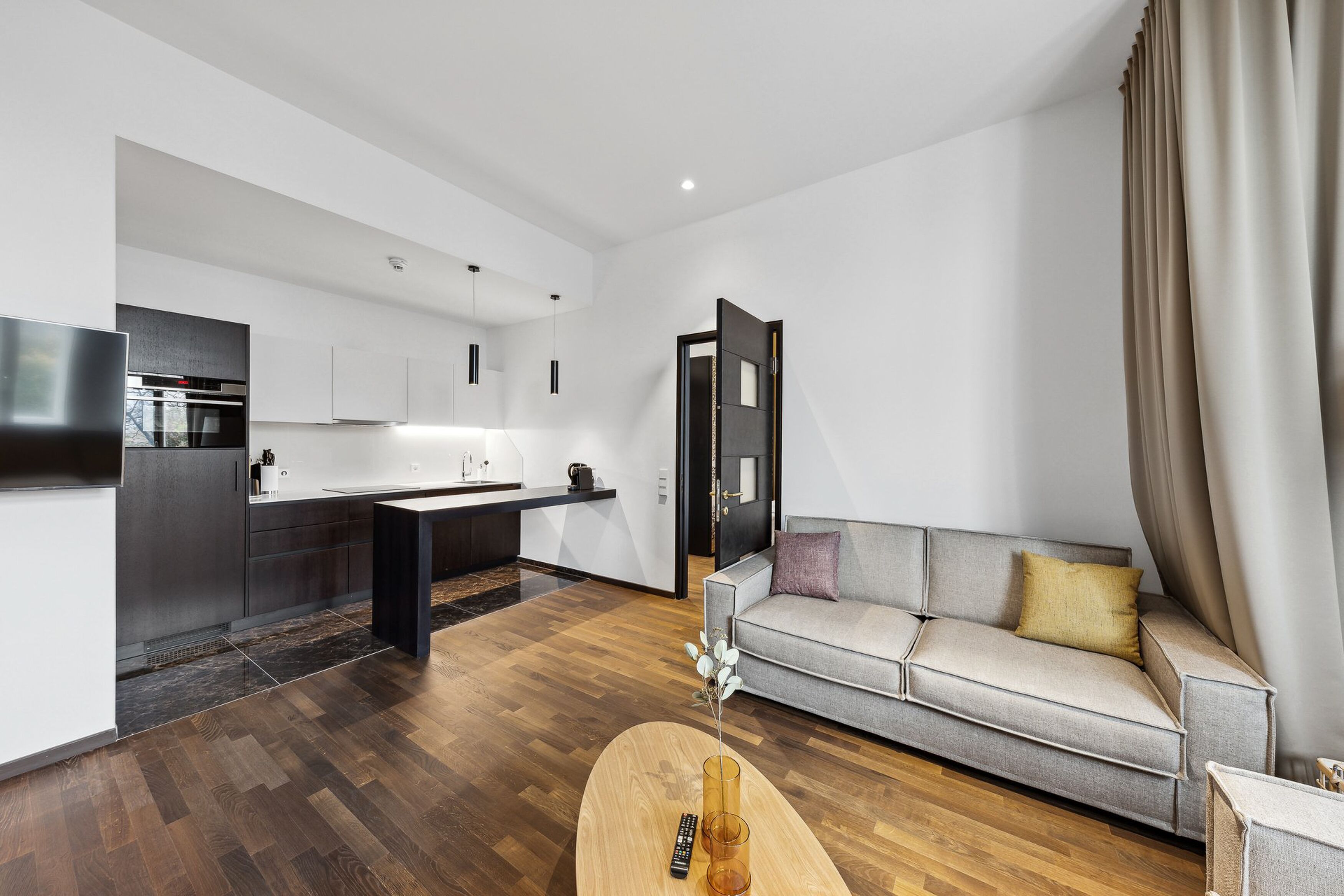 Uma sala de estar moderna com sofá, mesa de centro e área de cozinha com geladeira e armários.