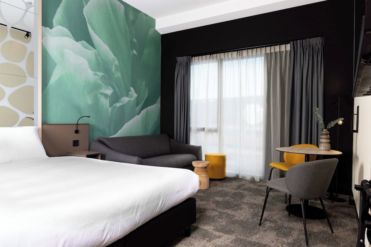Quarto de hotel com cama, sofá, mesa pequena, cadeira e um grande mural abstrato na parede.