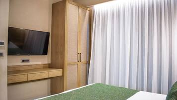 Quarto de hotel com cama, televisão presa a um móvel de madeira e janela com cortinas brancas.
