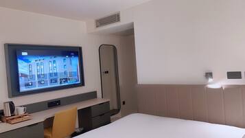 Quarto de hotel com cama, escrivaninha com computador, televisão fixada na parede e chuveiro.