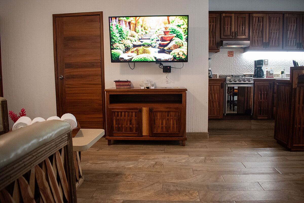 Uma sala de estar com um móvel de madeira para TV, uma televisão de tela plana fixada na parede, e uma cozinha com armários de madeira e fogão.