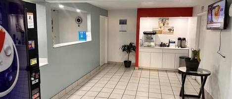 Um saguão de hotel com uma máquina de venda automática, uma pequena mesa com uma planta, uma máquina de café e uma televisão na parede.