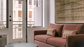 Uma sala de estar com sofá, uma mesa redonda, radiador e janela com vista para edifícios.