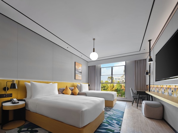 Quarto de hotel moderno com uma cama, duas mesas de cabeceira, uma escrivaninha, uma cadeira, uma televisão e uma grande janela com vista para edifícios e árvores.