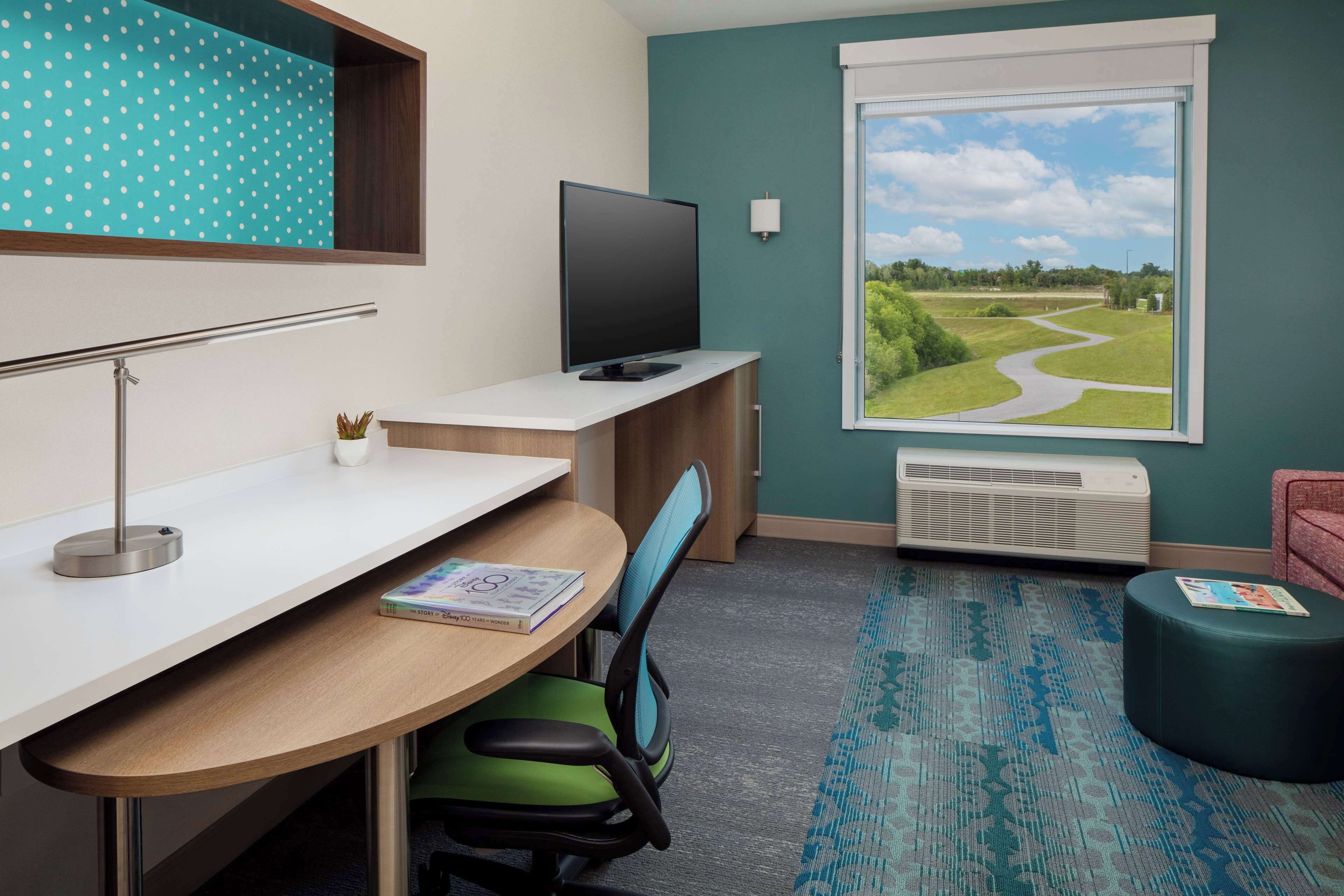 Quarto moderno com escrivaninha, cadeiras, televisão, ampla janela com vista para uma paisagem verde e tapete estampado.