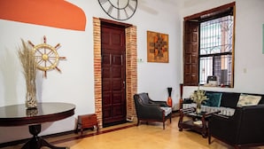 Uma sala de estar com uma mesa redonda, um sofá, uma poltrona, um relógio e uma porta.