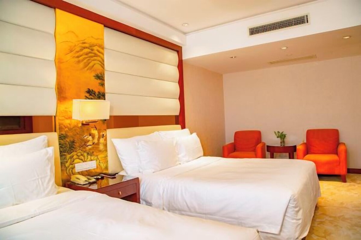 Quarto de hotel com duas camas, uma luminária na parede e duas cadeiras laranja.