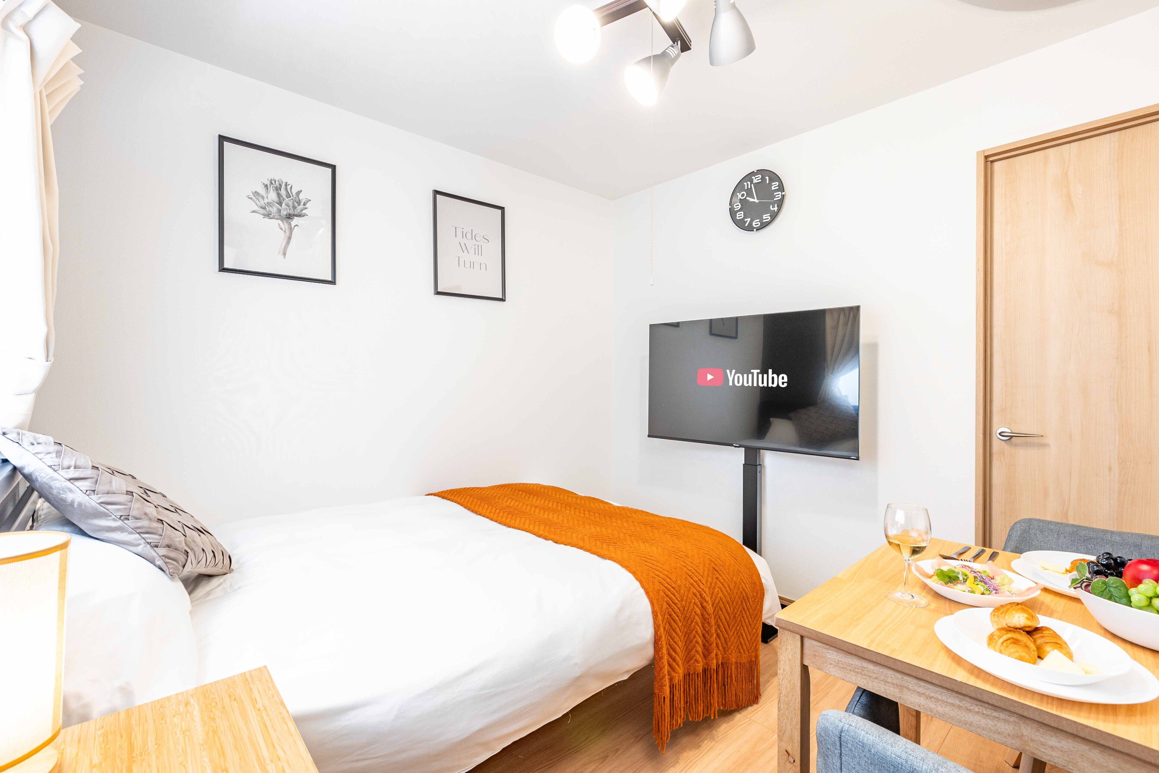 Quarto de hotel com cama, televisão na parede, mesa de jantar com pratos de comida e uma taça de vinho, e uma porta.
