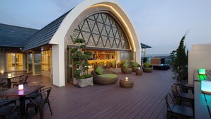 Uma área externa com mesas redondas e cadeiras acolchoadas, um bar com uma grande janela em arco e um deck de madeira.