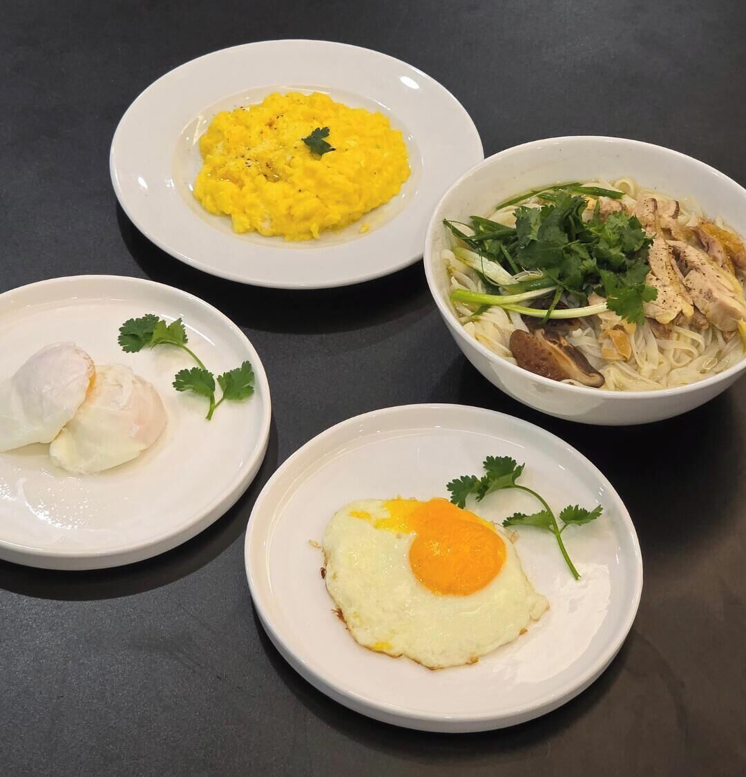 Uma mesa com quatro pratos de comida: ovos mexidos, uma tigela de macarrão com legumes, um prato de ovos pochê e um prato de arroz amarelo.