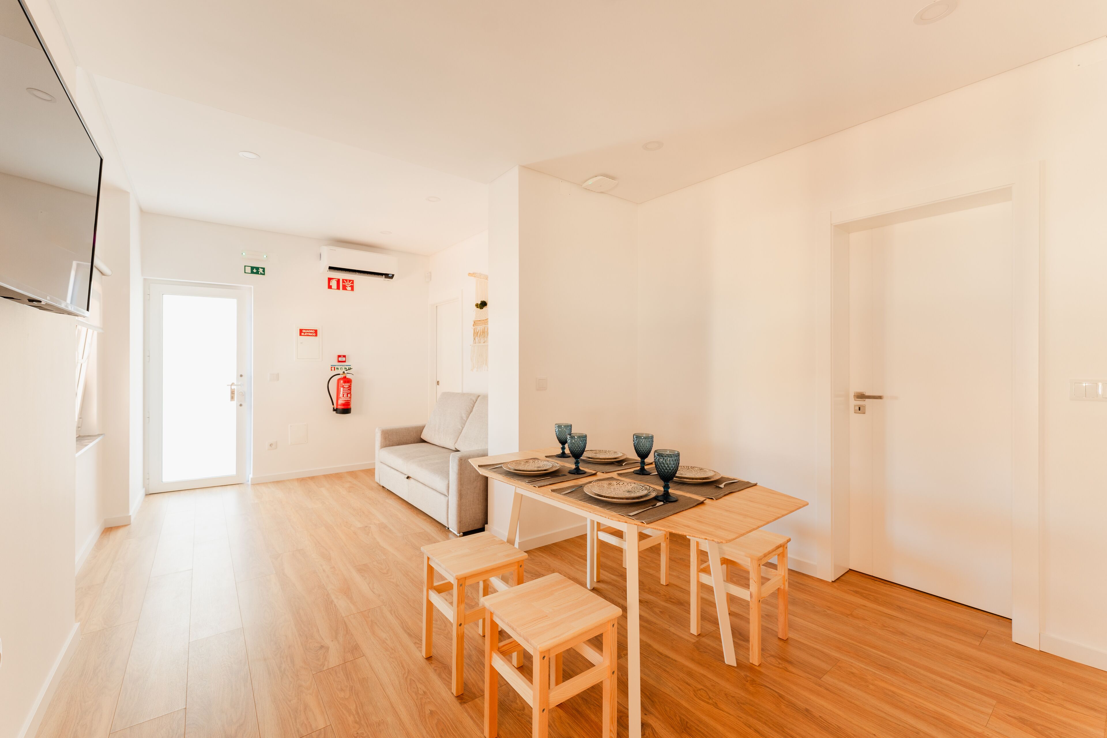 Uma sala de jantar luminosa e moderna, com uma mesa de madeira posta para quatro pessoas, um sofá e um extintor fixado na parede.